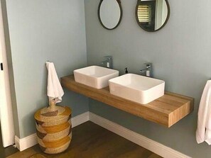 Um banheiro com duas pias brancas em uma bancada de madeira, um cesto redondo de vime e dois espelhos redondos na parede.