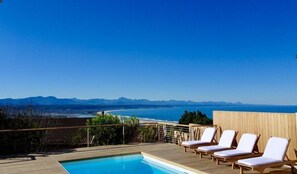 Uma área à beira da piscina com espreguiçadeiras e vista para o oceano e montanhas.
