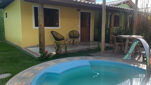Área de piscina com uma casa amarela, duas cadeiras e uma mesa.