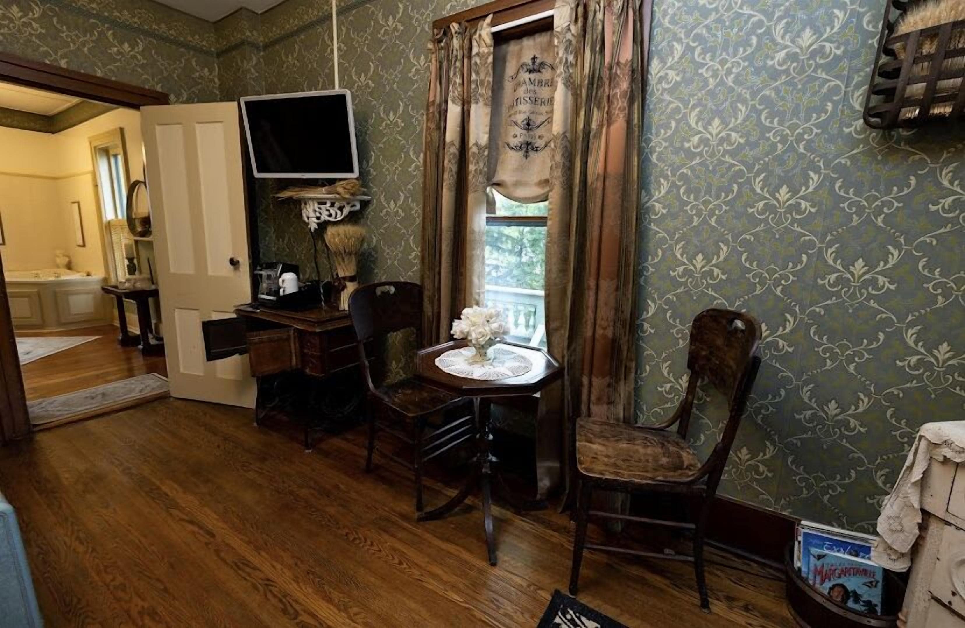Um quarto com piso de madeira, uma mesa de jantar com cadeiras, uma televisão em um móvel e uma janela com cortinas.