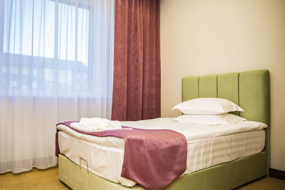 Uma cama bem arrumada com cabeceira verde, travesseiros brancos e manta roxa num quarto com soalho de madeira e cortinas vermelhas.