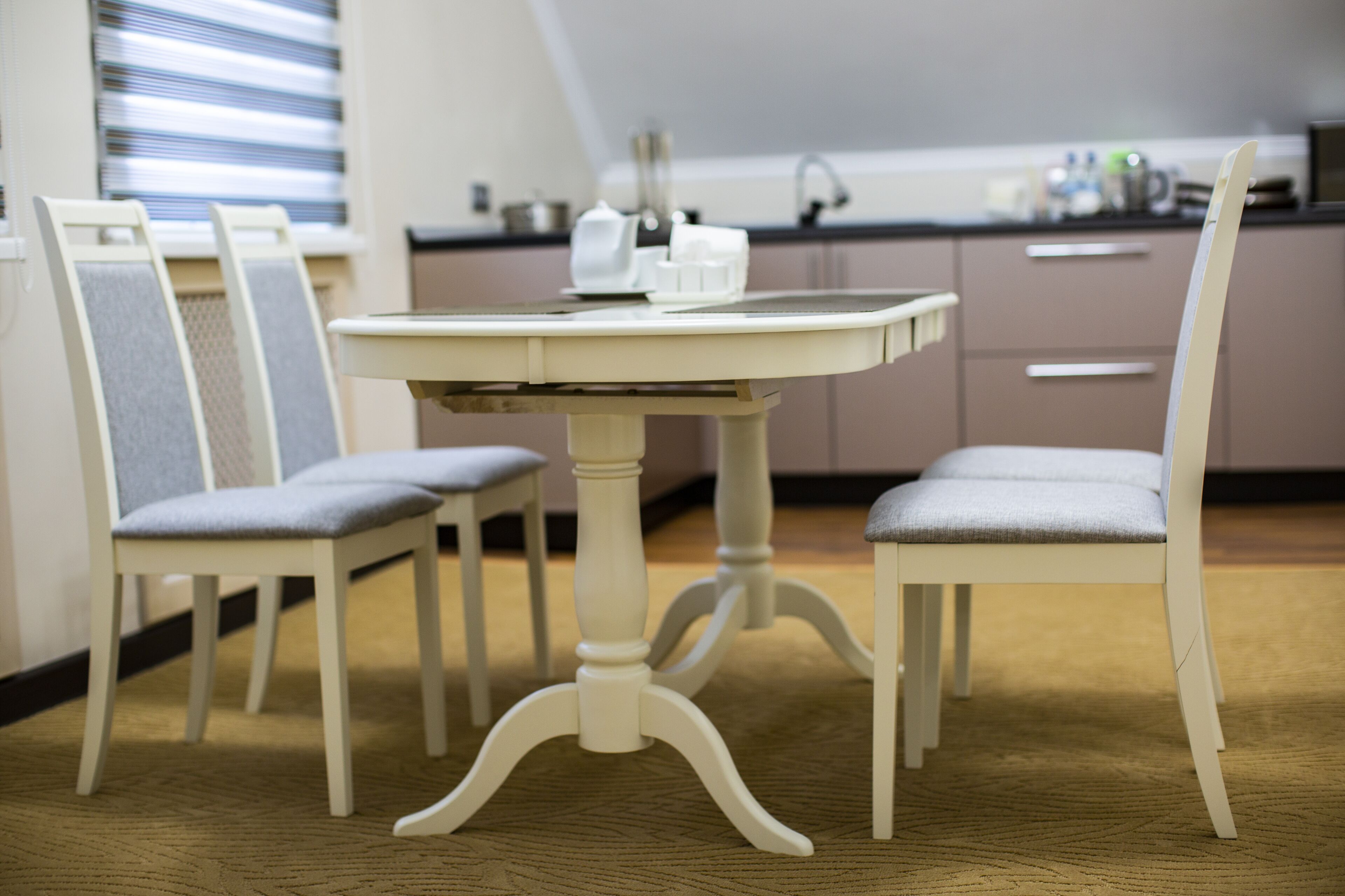 Uma área de refeições com uma mesa e cadeiras brancas, uma cozinha ao fundo e uma janela com persianas.