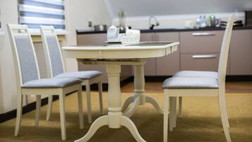 Uma área de refeições com uma mesa e cadeiras brancas, uma cozinha ao fundo e uma janela com persianas.