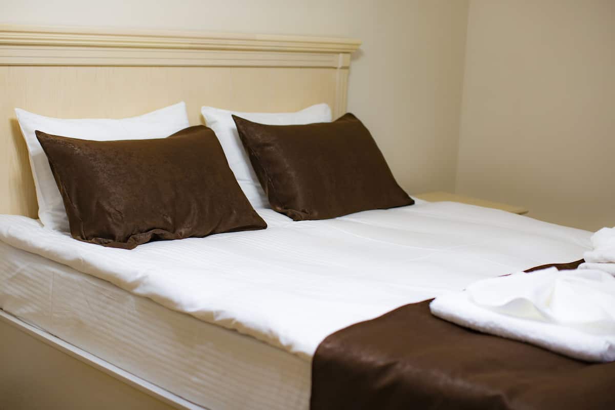 Cama bem arrumada com roupa de cama branca e marrom, cabeceira e parede lisa.