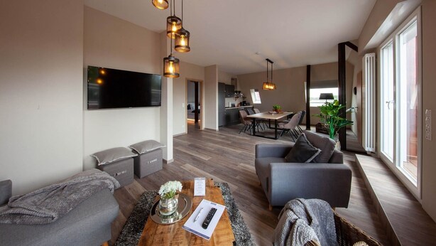 Uma sala de estar moderna com uma televisão de tela plana, sofás cinzas, uma mesa de centro de madeira com um vaso e controle remoto, e um espaço de jantar com mesa e cadeiras.