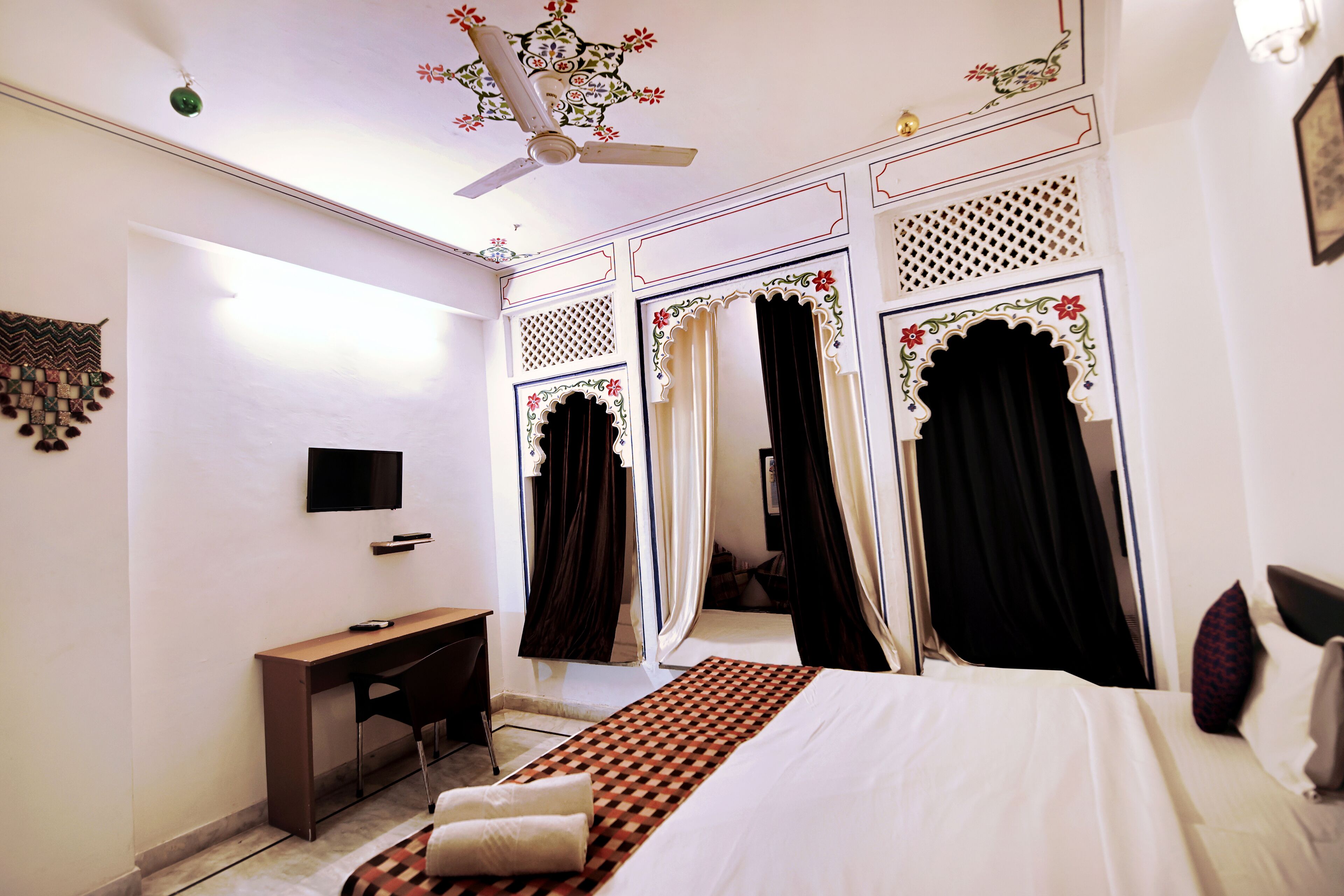 Quarto de hotel com cama, escrivaninha, televisão e ventilador de teto.