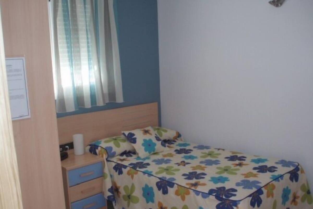 Quarto com uma cama de solteiro, cômoda de madeira com gavetas, janela com cortinas e luminária fixada na parede.
