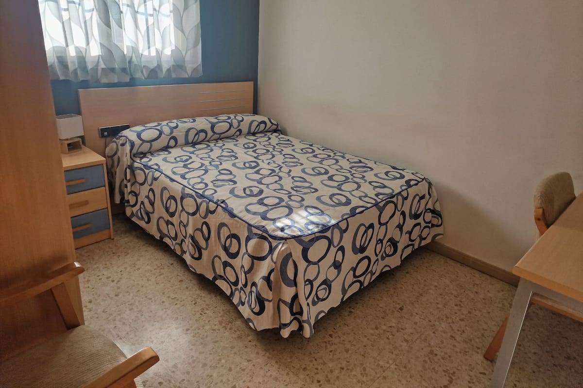 Quarto com cama, mesa de cabeceira de madeira, uma cadeira e janela com cortinas leves.