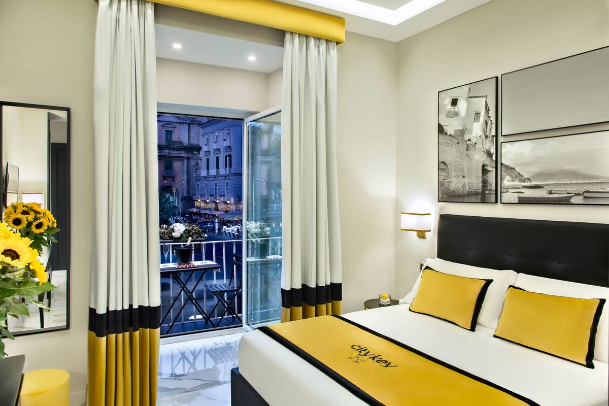 Quarto de hotel com cama, cortinas, varanda com mesa e cadeiras, e obras de arte na parede.