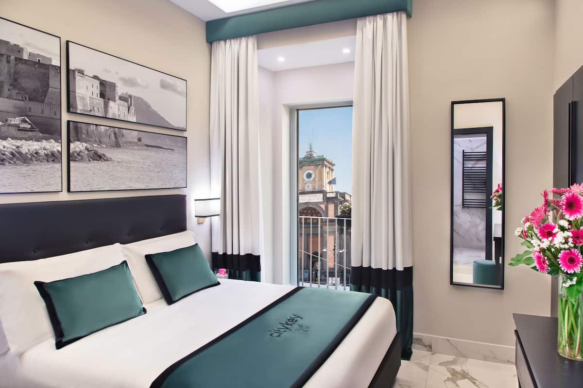 Quarto de hotel com uma cama grande, vista para um campanário e um vaso de flores em uma mesinha de cabeceira.