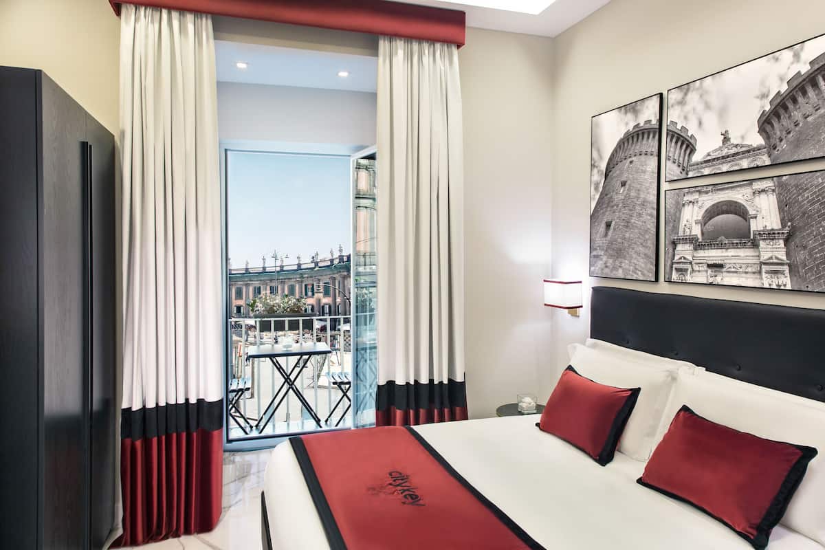 Quarto de hotel com cama, cortinas, varanda e vista para edifícios, além de um quadro na parede.