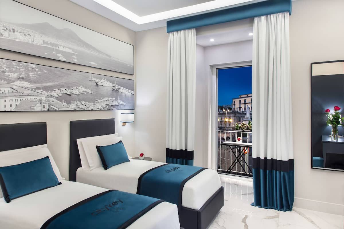 Quarto de hotel com duas camas, varanda com guarda-corpo e vista para a cidade.