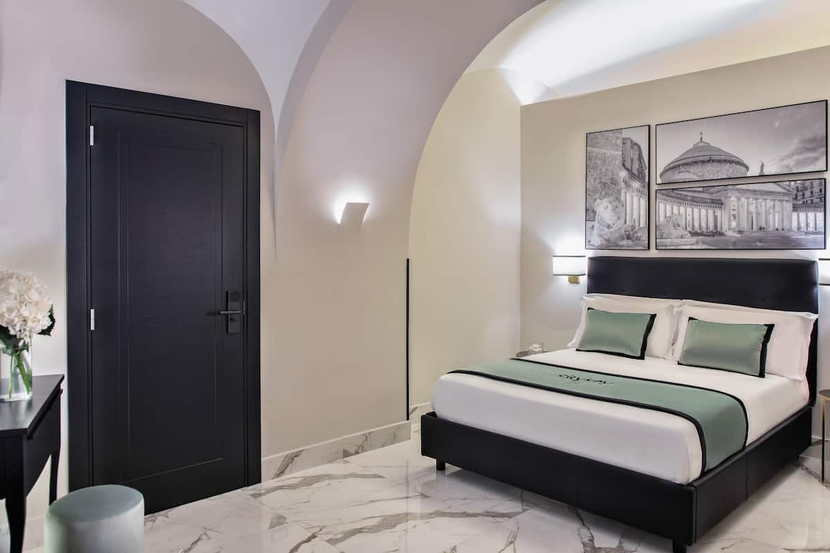 Quarto moderno com piso de mármore, porta escura, cama com roupa de cama verde e branca e luminária fixada na parede.