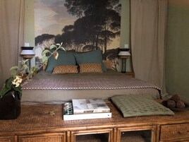 Quarto com cama, duas luminárias de cabeceira, mesa de cabeceira com livros e uma tigela decorativa, e um grande mural na parede.