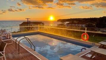 Piscina com espreguiçadeiras e boia, com vista para o mar ao pôr do sol.