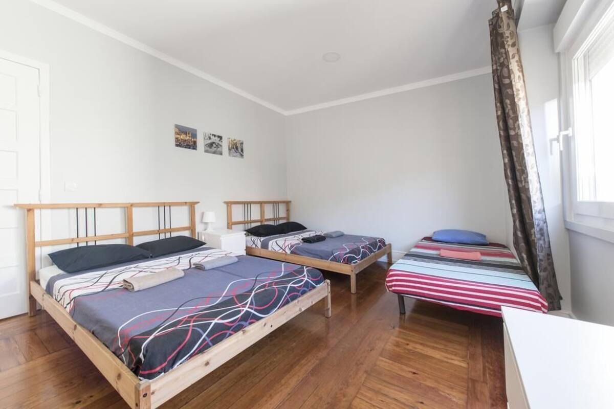 Quarto pequeno e limpo, com duas camas de madeira, uma escrivaninha branca e uma janela com cortinas.