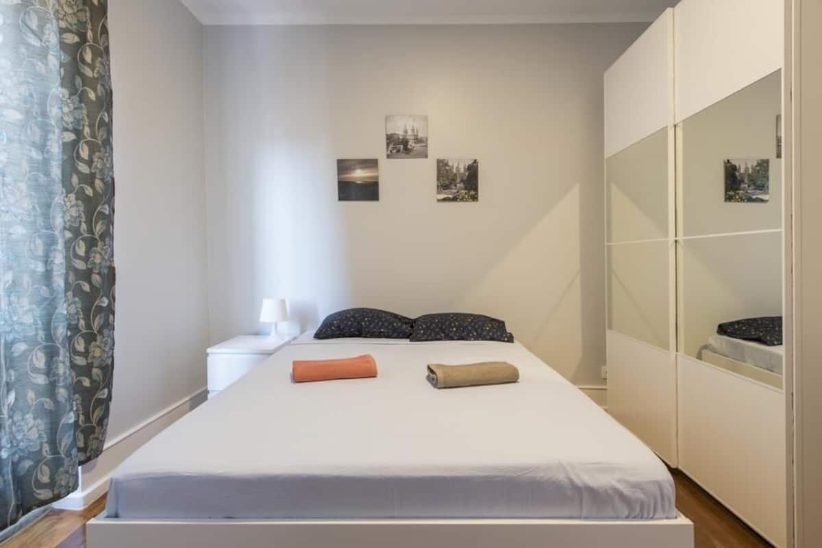 Cama bem arrumada com duas toalhas enroladas, mesa de cabeceira com abajur e guarda-roupa com espelho.