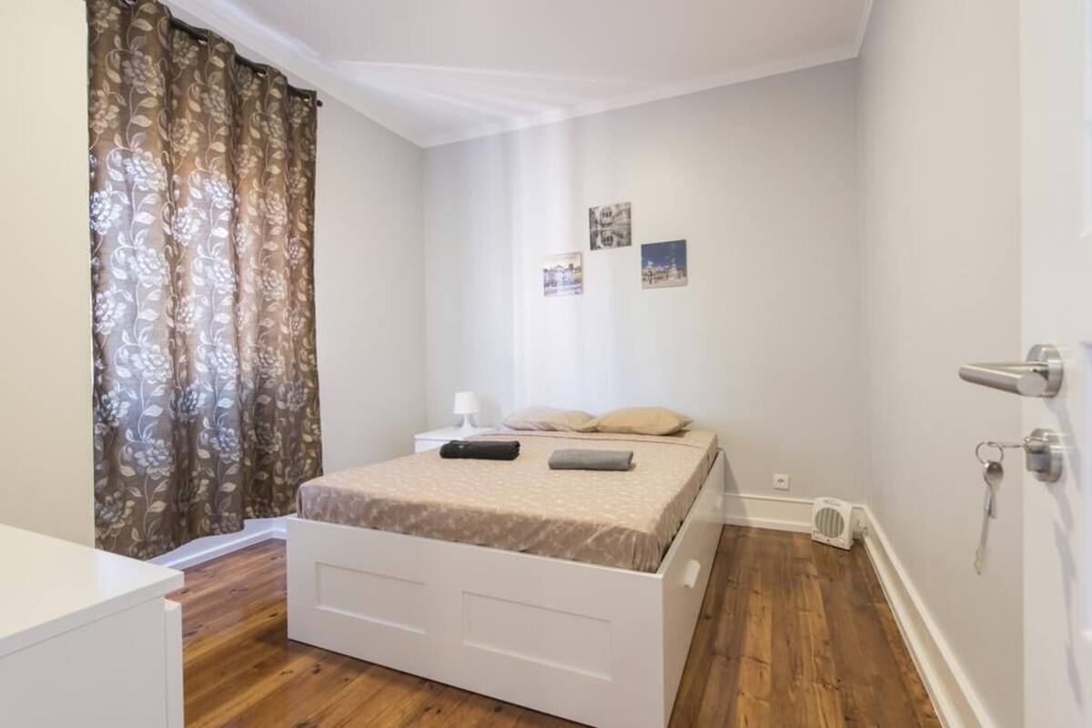 Quarto com cama, mesa de cabeceira, luminária e janela com cortinas.