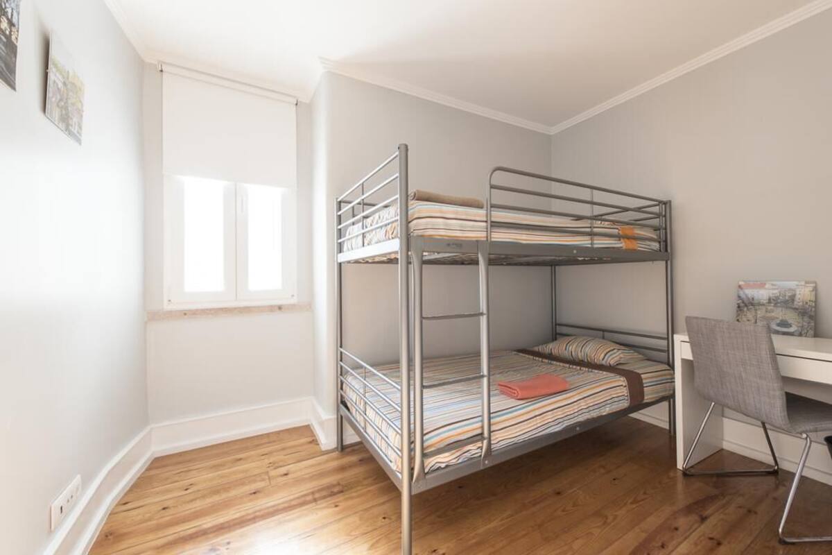 Quarto com beliche, escrivaninha e cadeira, piso de madeira e uma janela.