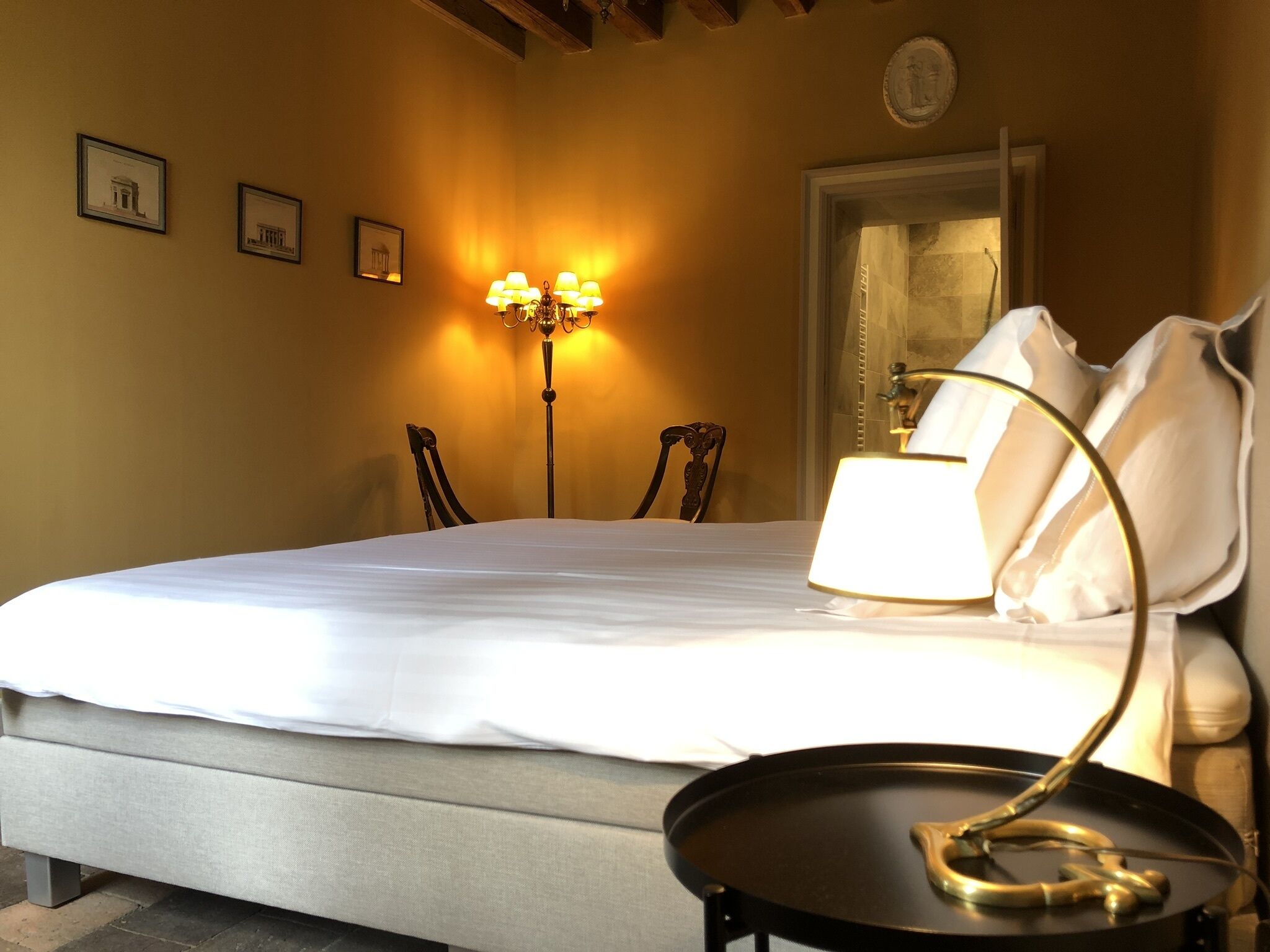 Cama bem arrumada com edredom branco e travesseiros, mesa de cabeceira com abajur, e quadros nas paredes.