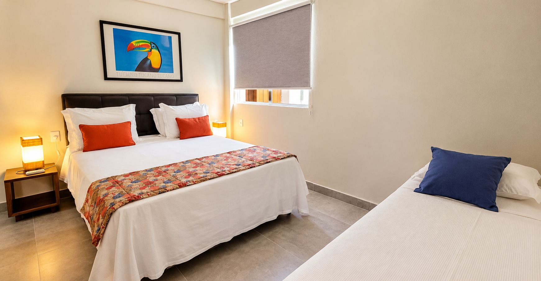 Quarto de hotel com uma cama grande, uma cama menor, uma mesa de cabeceira com abajur, uma janela com persiana e uma moldura com um quadro na parede.