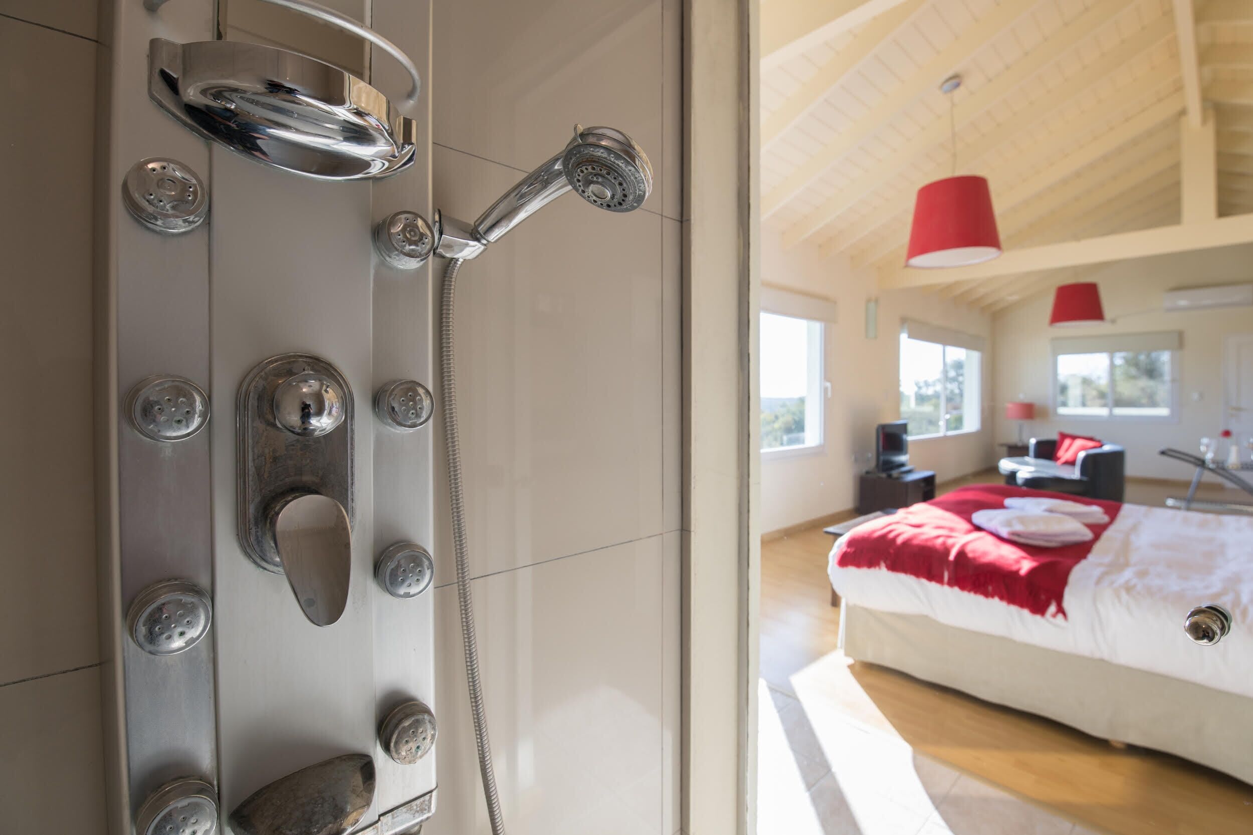 Casa de banho moderna com chuveiro, cama com manta vermelha e quarto com teto de madeira e candeeiros pendentes vermelhos.