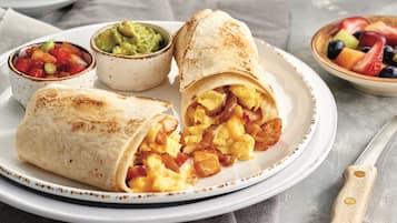 Um prato com um burrito, guacamole e uma saladeira de frutas.