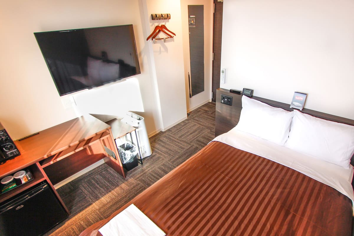 Quarto de hotel com cama, escrivaninha, televisão, lixeira e cabideiro.