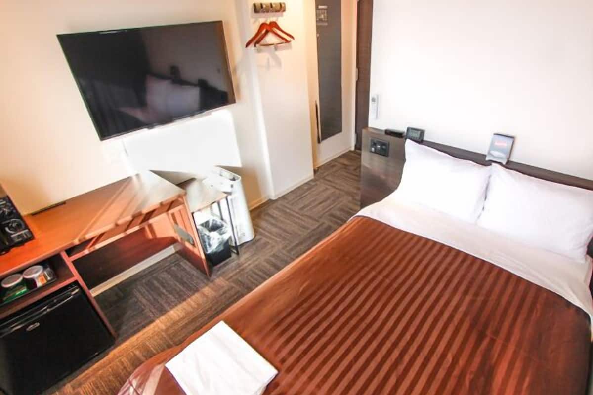 Quarto de hotel com uma cama, televisão na parede, escrivaninha com cadeira e uma pequena lixeira.