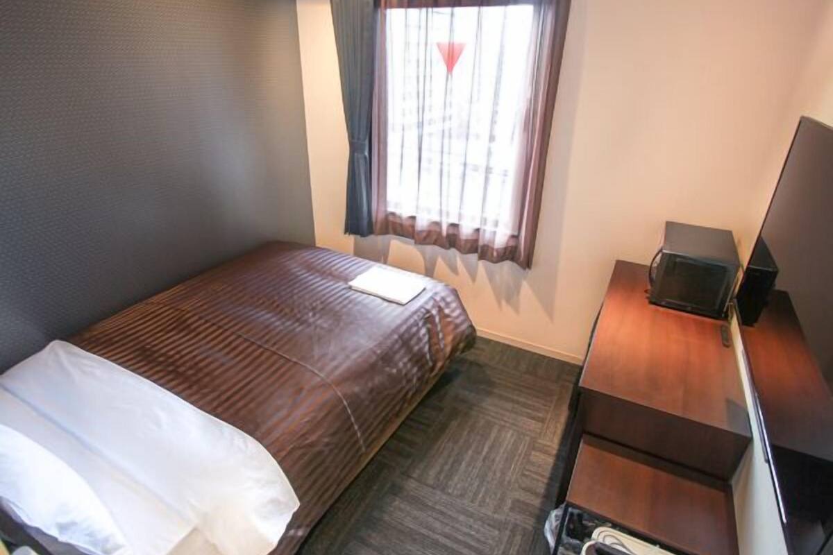 Quarto de hotel pequeno e bem organizado, com uma cama de solteiro, uma escrivaninha com computador e uma janela com cortinas.