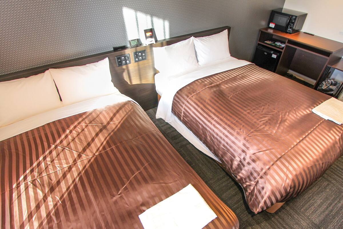 Duas camas com roupa de cama listrada em tons de castanho, uma mesa de cabeceira com rel ógio e uma pequena escrivaninha com micro-ondas.