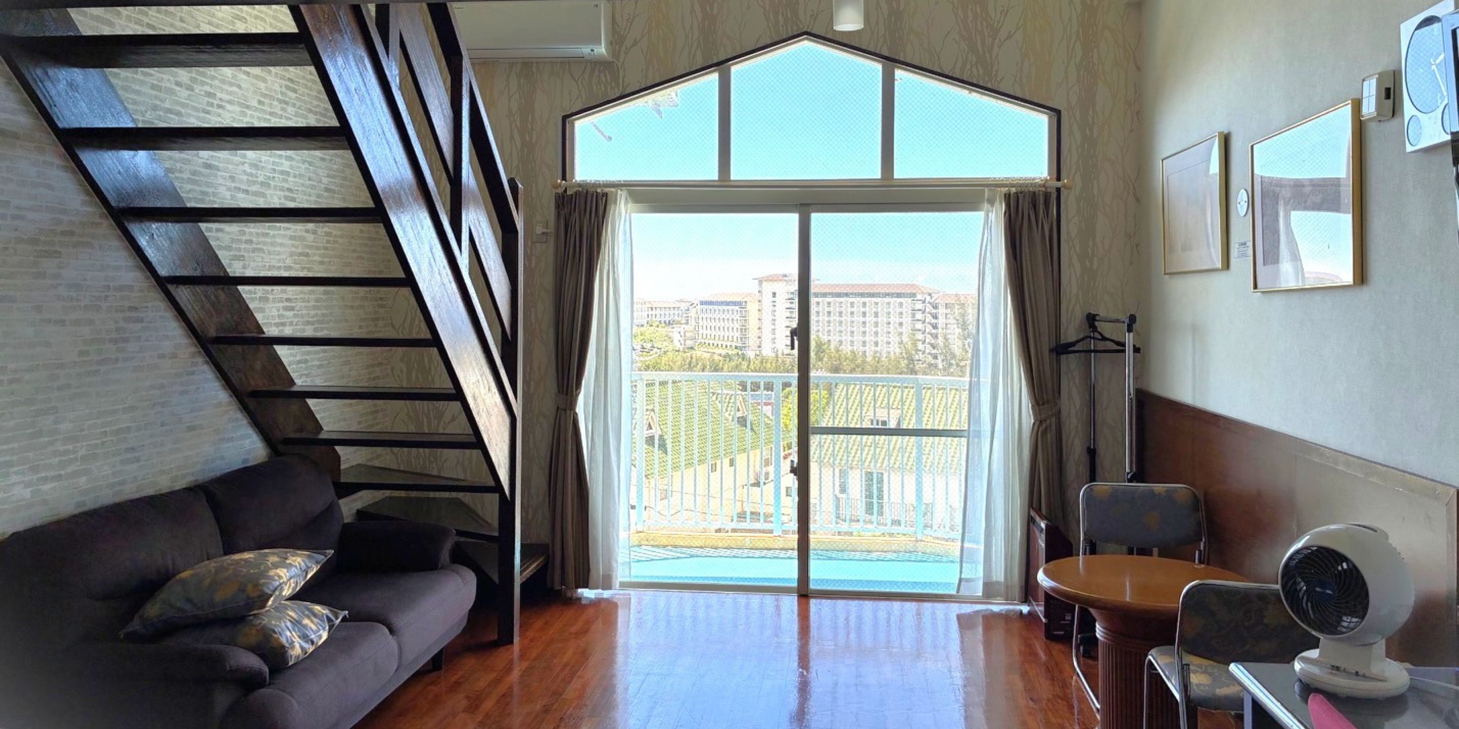 Quarto com sofá, mesa, ventilador e vista para edifícios através de uma grande janela.