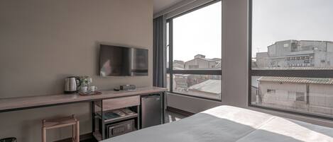 Quarto de hotel moderno com uma cama grande, uma mesa de trabalho com televisão de tela plana, cafeteira e vista para prédios por grandes janelas.