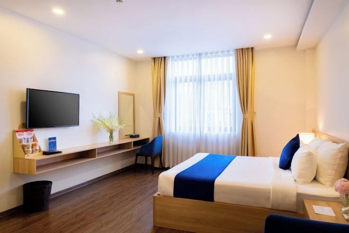 Quarto de hotel com cama, escrivaninha com cadeira, televisão e janela com cortinas.