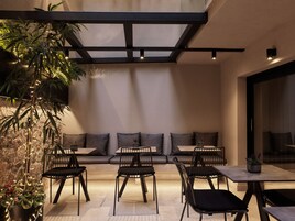 Uma área de estar interna com sofá, cadeiras e uma mesa, cercada por plantas e uma estrutura de teto de madeira.