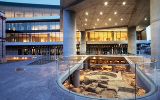 Um edifício moderno com átrio de vidro, pátio central com vestígios arqueológicos e mistura de arquitetura contemporânea e clássica.