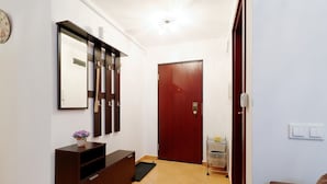 Um corredor com porta de madeira, espelho, mesa pequena com vaso e lixeira.