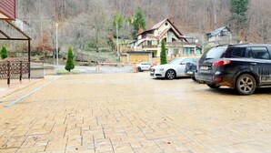 Uma área de estacionamento pavimentada com dois carros estacionados, um edifício com telhado vermelho e um morro arborizado ao fundo.