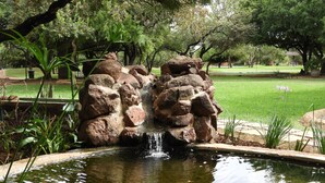 Um jardim com uma pequena cascata e um lago rodeado por rochas e vegetação.