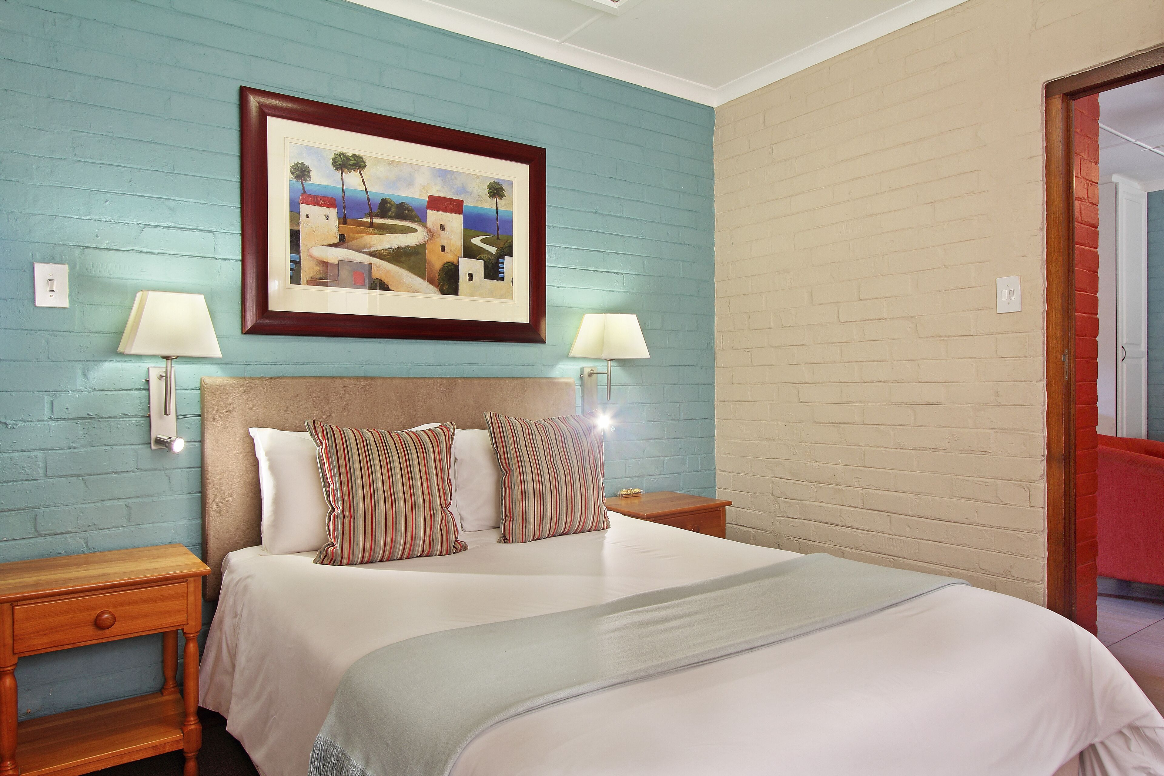 Quarto de hotel com cama, duas luminárias de cabeceira, mesa de apoio, um quadro na parede e uma porta de madeira.