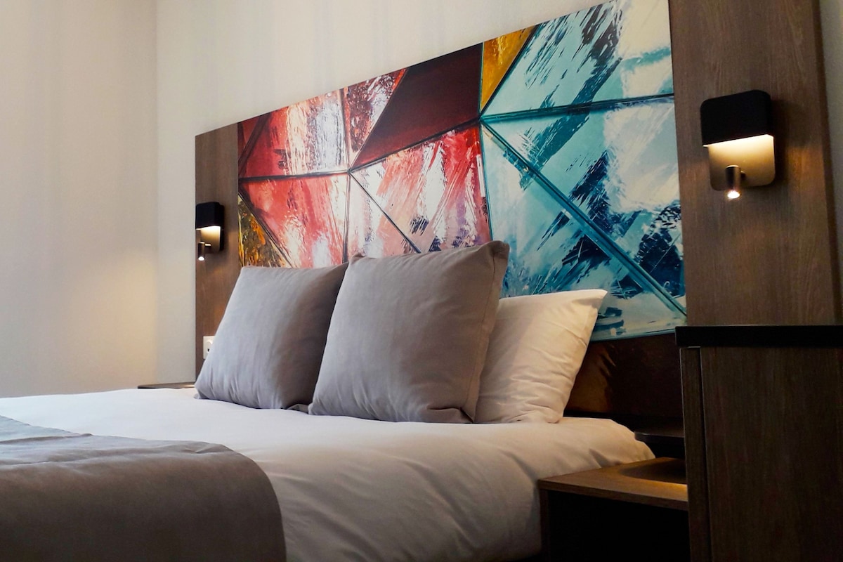 Quarto de hotel com uma cama grande, um quadro abstrato colorido na parede e duas luminárias fixadas na parede.
