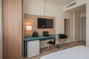 Quarto de hotel com escrivaninha azul-esverdeada, televisão de tela plana, mini-geladeira, cadeira e cama.