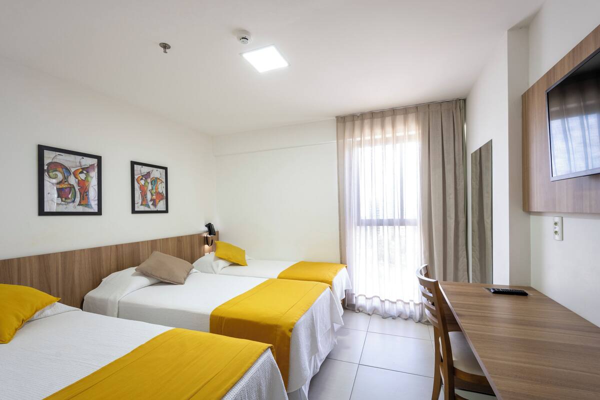 Quarto de hotel com duas camas, uma escrivaninha de madeira, televisão e janela com cortinas.
