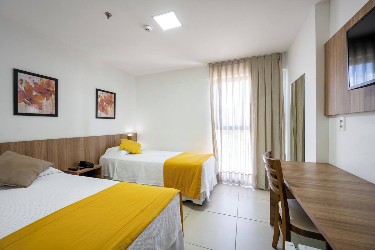 Quarto de hotel com duas camas, uma escrivaninha de madeira e uma televisão na parede.