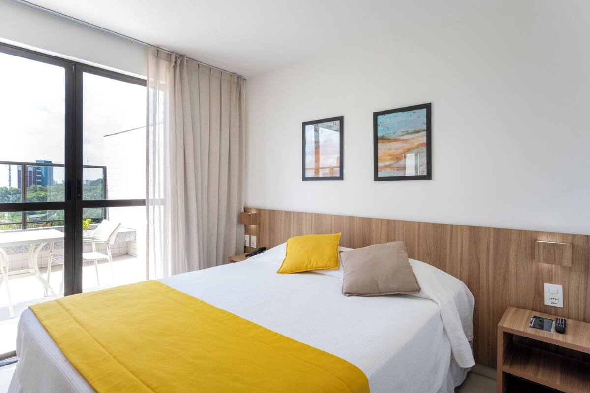 Quarto de hotel com uma cama grande, uma varanda com mesa e cadeiras, e duas fotos emolduradas na parede.