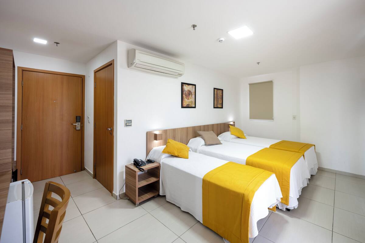 Quarto de hotel com duas camas, uma escrivaninha, uma cadeira e um ar condicionado.