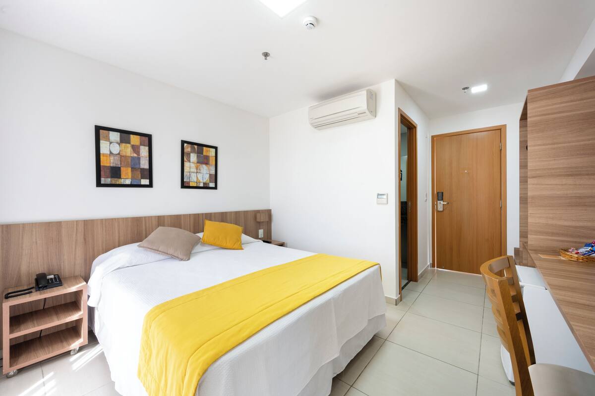 Quarto de hotel moderno com uma cama grande, uma mesinha de cabeceira com telefone, uma escrivaninha com cadeira e um guarda-roupa.