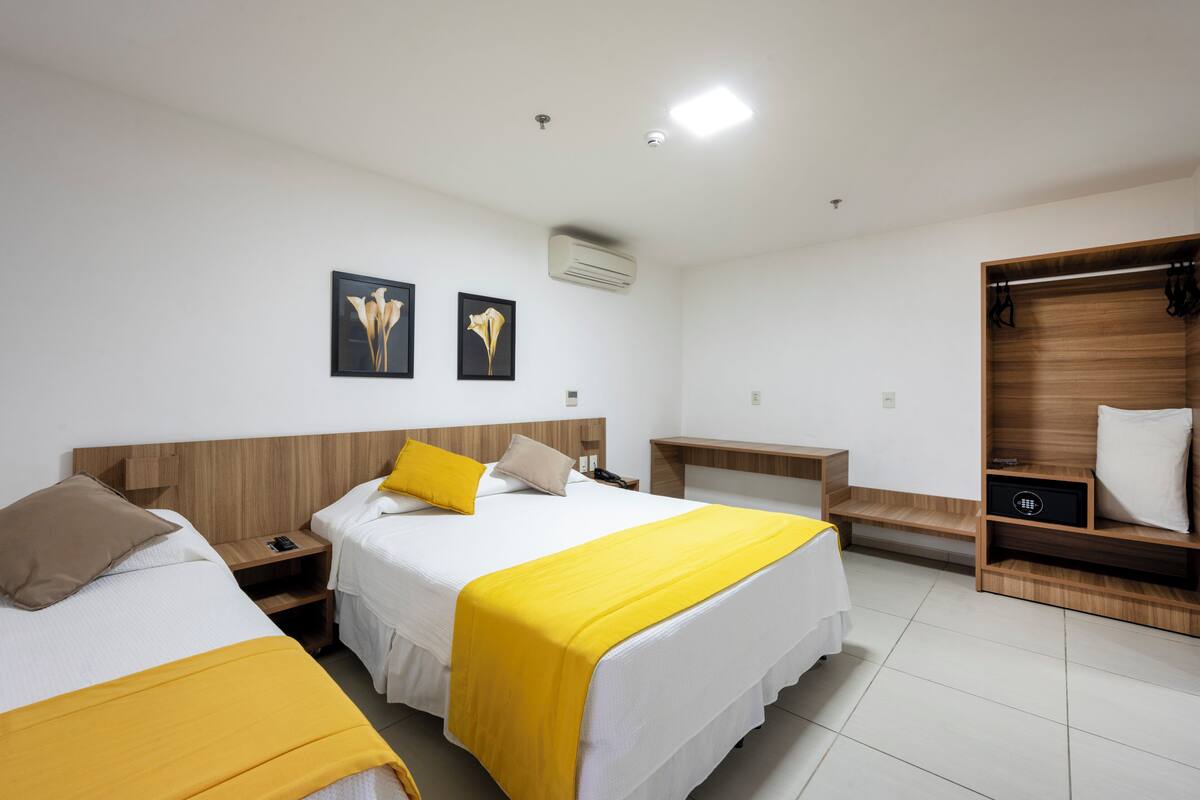 Quarto de hotel com duas camas, guarda-roupa de madeira, mesa de cabeceira e ar condicionado fixado na parede.