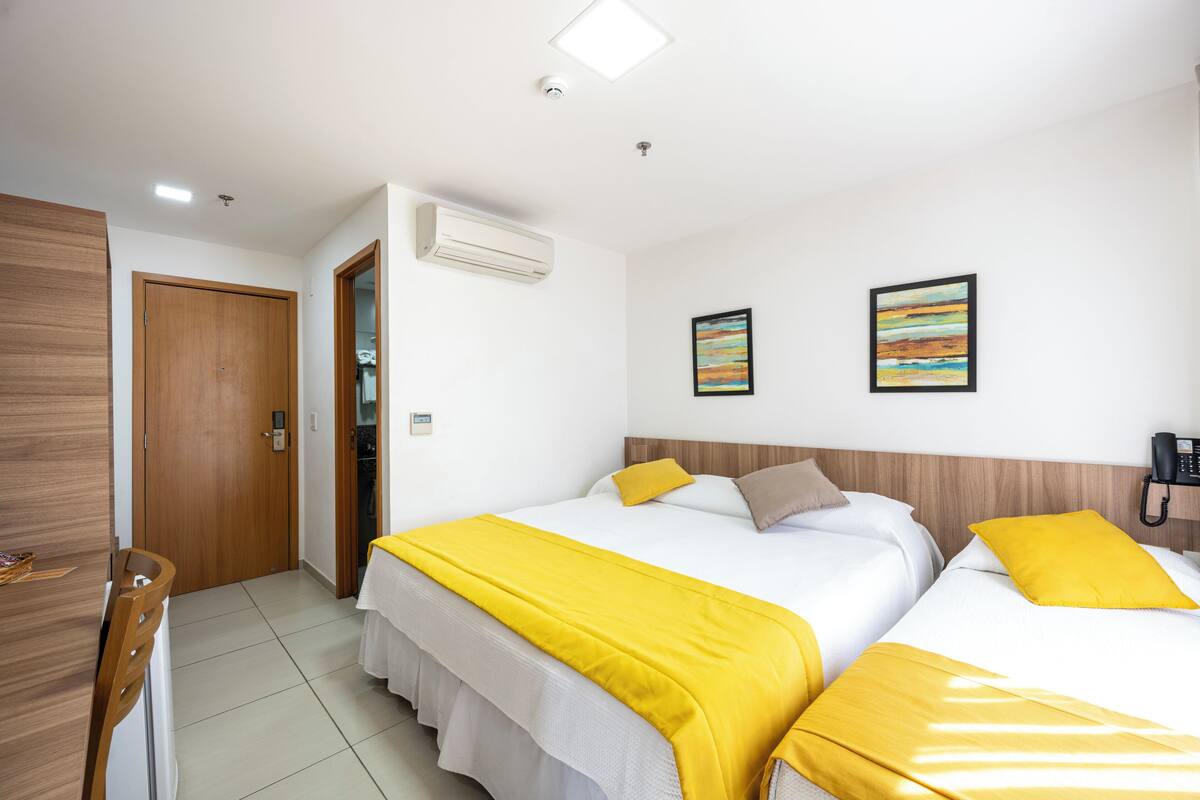 Quarto de hotel com duas camas, um guarda-roupa de madeira, uma escrivaninha e um telefone.