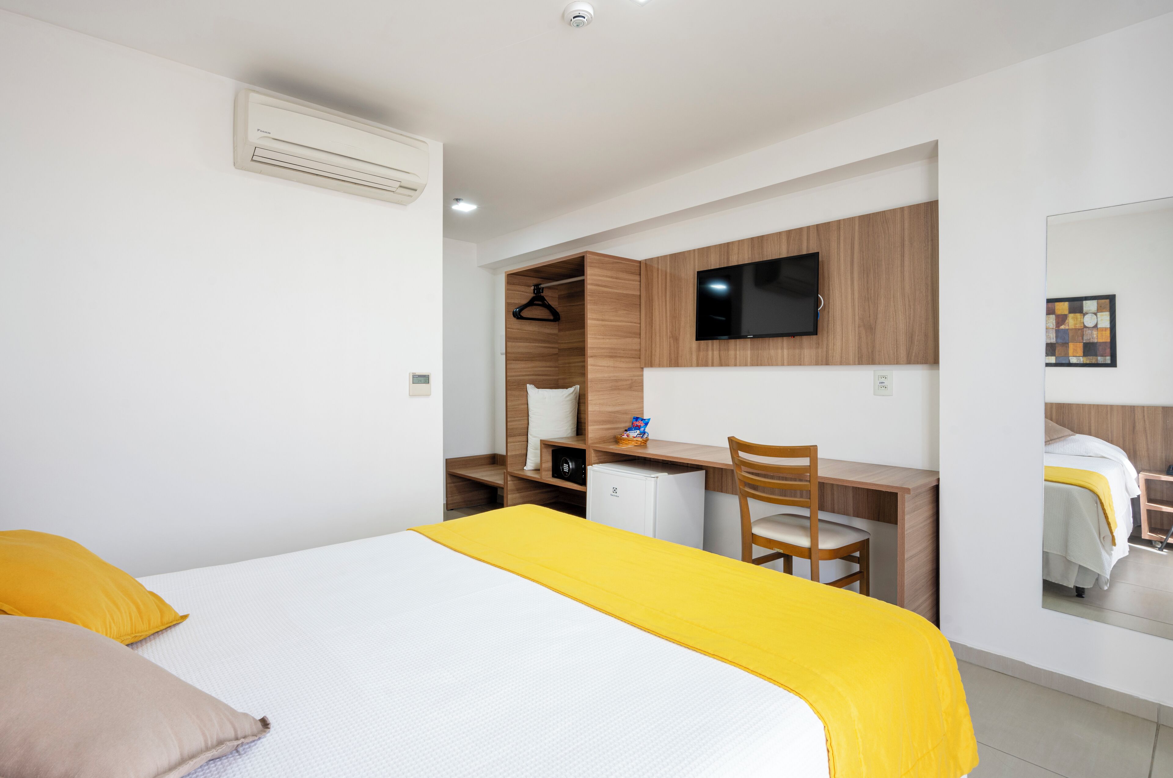 Quarto de hotel moderno com cama, escrivaninha, cadeira, televisão e um pequeno frigorífico.